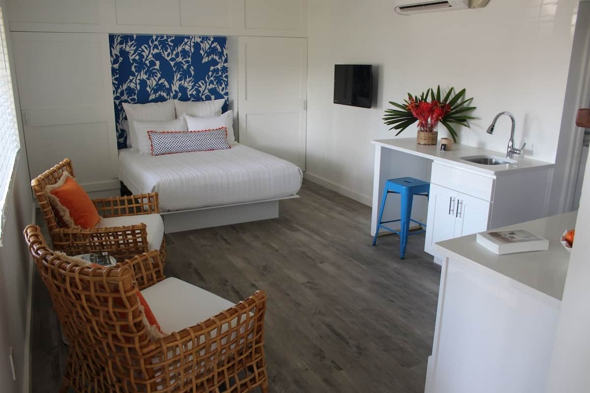 Quarto compacto com cama, uma pequena escrivaninha, uma cadeira, pia e um vaso de planta.