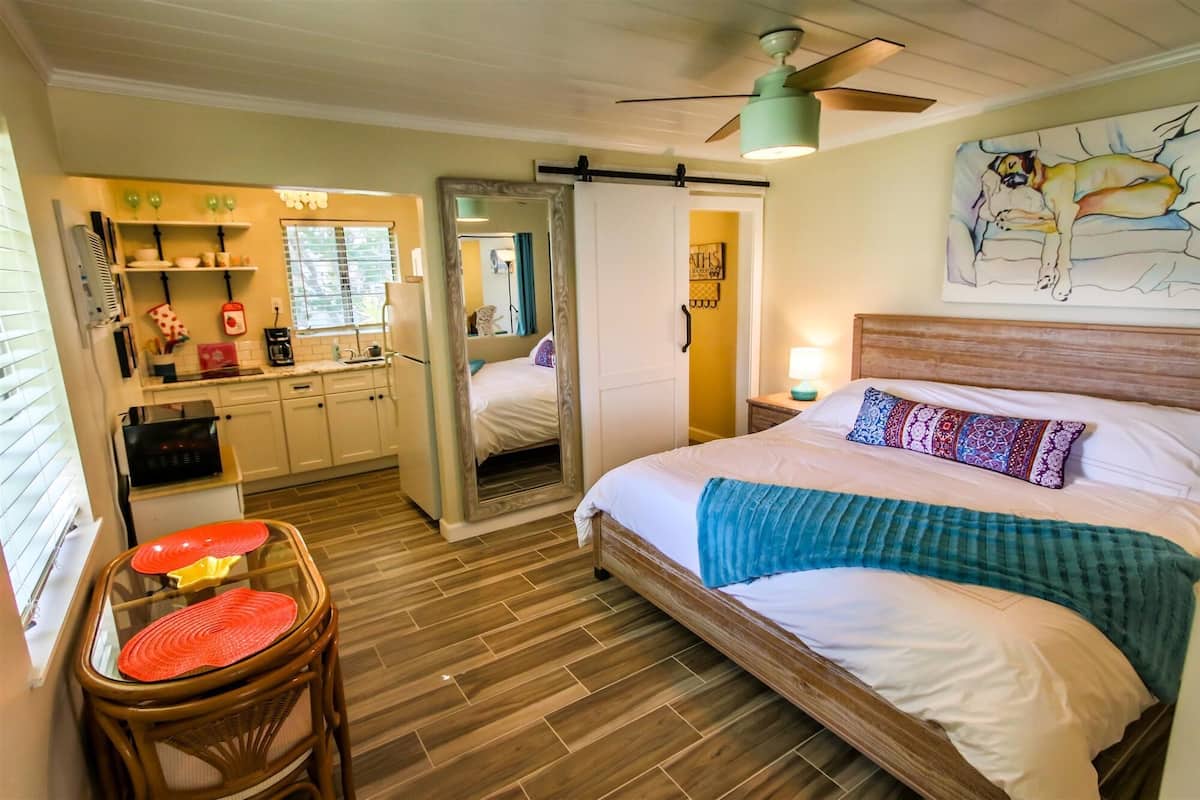 Quarto com cama, mesa de cabeceira, abajur, ventilador de teto, banheiro com chuveiro e cozinha equipada com geladeira e micro-ondas.