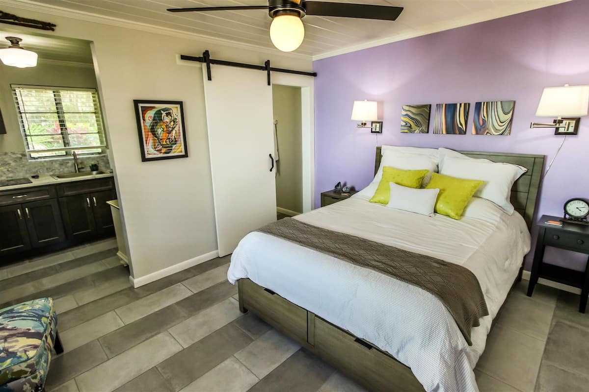 Quarto com cama, luminárias de cabeceira, mesa de cabeceira, ventilador de teto, janela com persianas, obra de arte emoldurada e banheiro com pia e espelho.