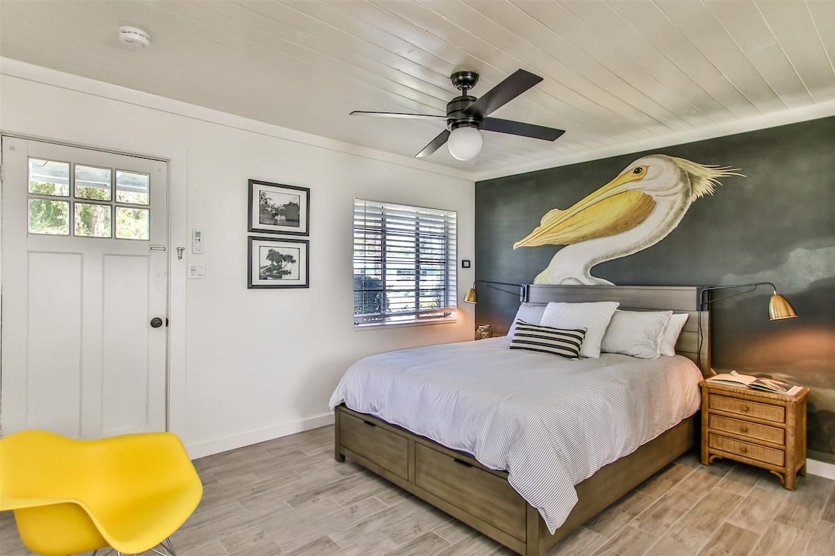 Quarto com cama, ventoinha no teto, janela com persianas, luminária na parede e um mural grande com gaivotas.