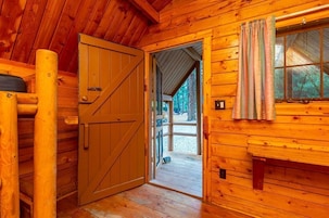 Interior de uma cabana de madeira com uma porta grande, uma janela com cortinas e um banco de madeira.