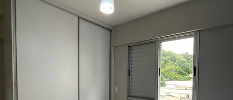 Quarto com cama, ventilador de teto, janela com persianas e vista para o verde lá fora.