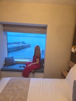 Quarto com cama, um assento junto à janela com vista para o mar e uma mesa de cabeceira com luminária.