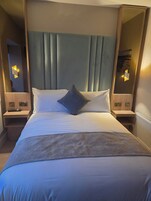 Uma cama bem arrumada com cabeceira, mesas de cabeceira com abajures e um espelho.