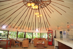 Quarto com teto alto sustentado por vigas de madeira, área de estar com móveis de vime e janela com vista para o verde lá fora.
