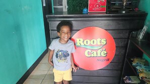 Uma criança está em pé num café, com um letreiro que diz 'Roots Café' e um aviso de distanciamento social no chão.