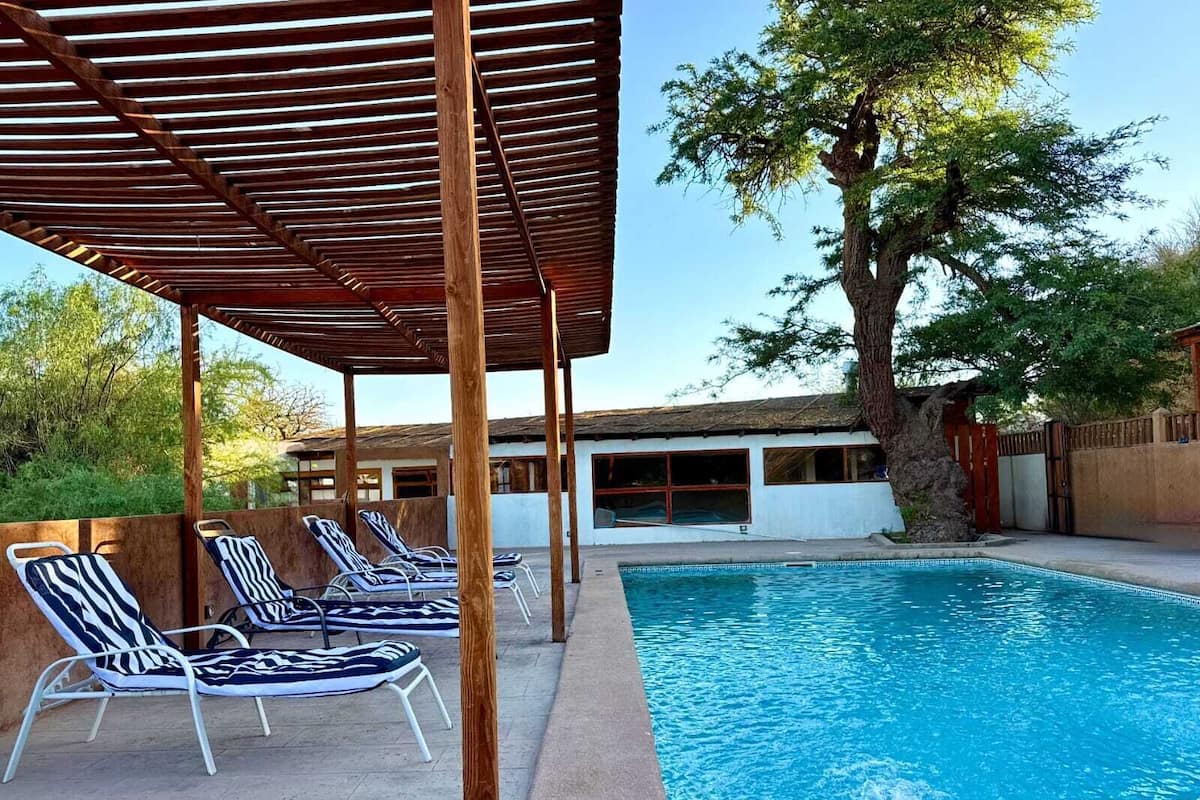 Piscina com espreguiçadeiras, um pergolado de madeira e um edifício ao fundo.