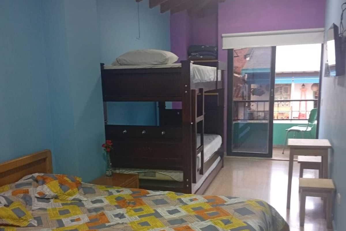 Quarto com beliches, cama com manta estampada, uma mesinha e vista através de uma porta.