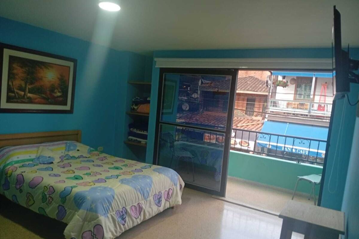 Quarto com cama, escrivaninha, cadeira, janela com vista para edifícios e um quadro na parede.