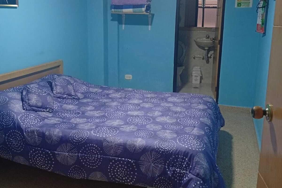 Quarto com cama, prateleira pequena, porta para banheiro e um quadro na parede.