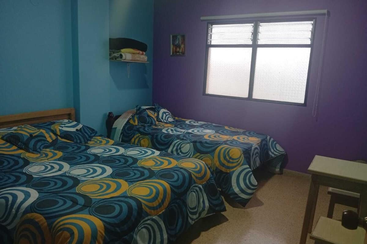Quarto com duas camas, uma janela, uma escrivaninha e uma prateleira com roupas dobradas.