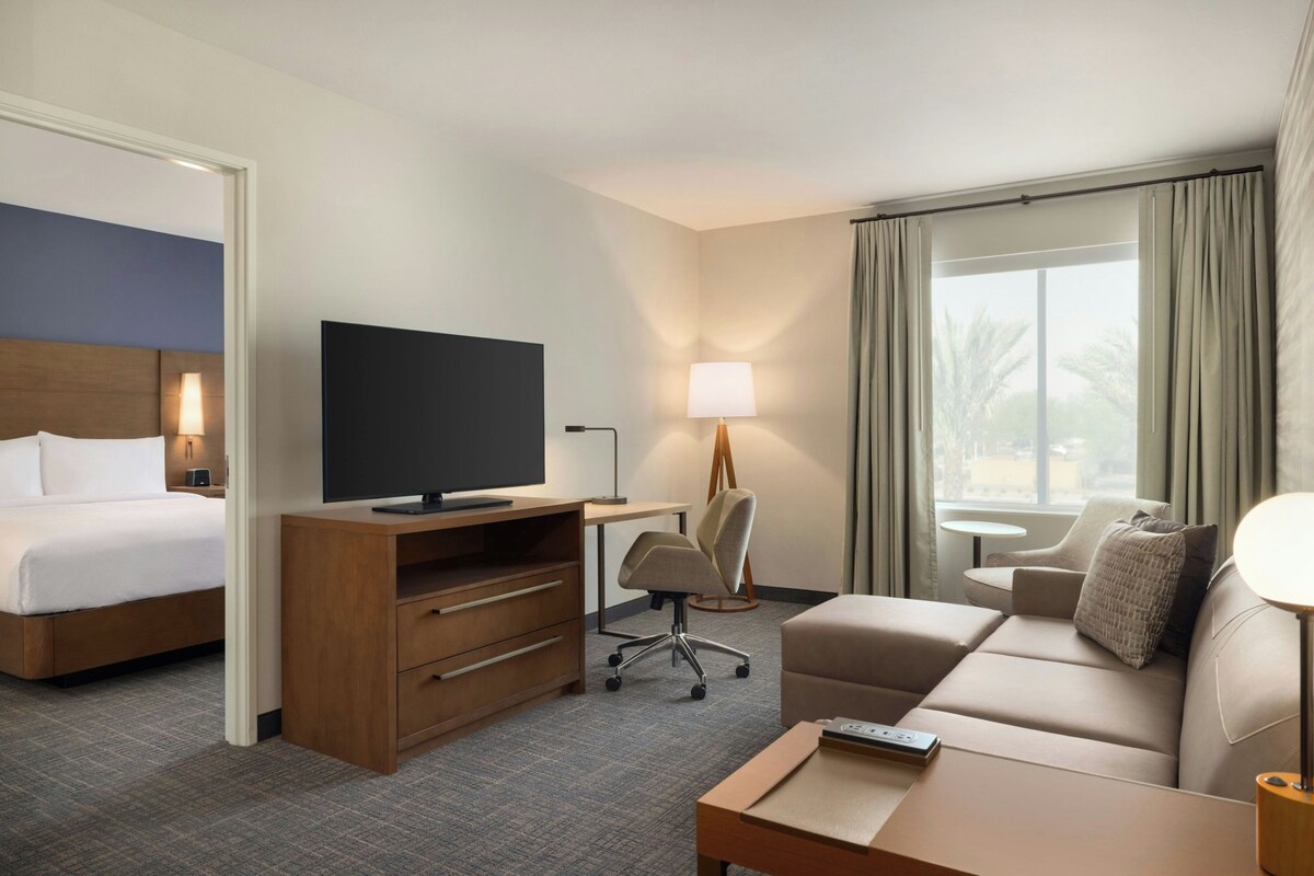 Quarto de hotel com cama, escrivaninha com televisão, cadeira, sofá e janela com cortinas.
