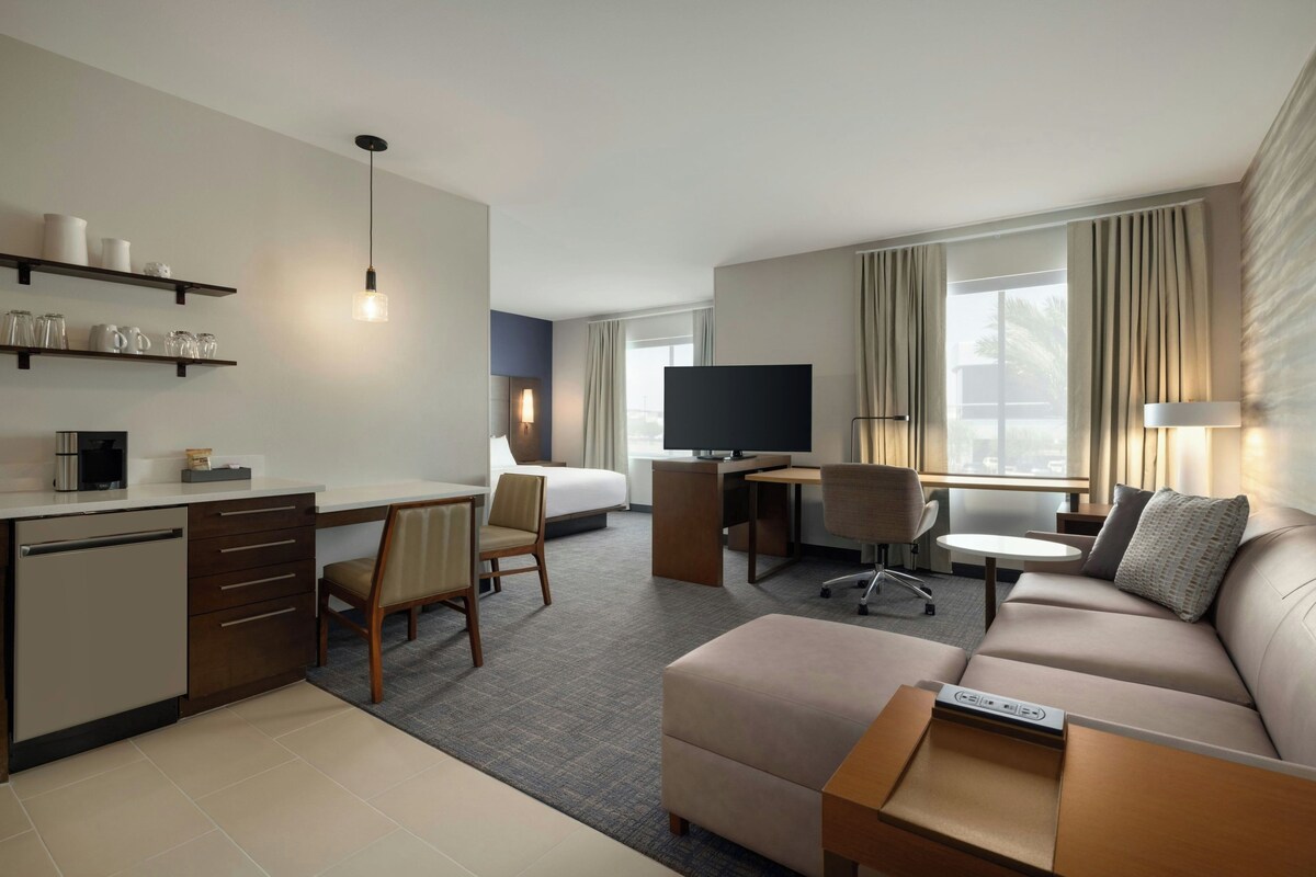 Quarto de hotel moderno com cozinha compacta, TV de tela plana, sofá, escrivaninha e área de refeições.