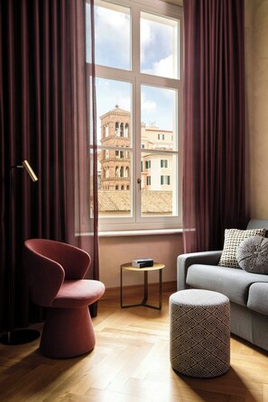 Quarto com vista para um edifício através de uma janela, uma cadeira vermelha, um sofá cinzento e um banquinho estampado.
