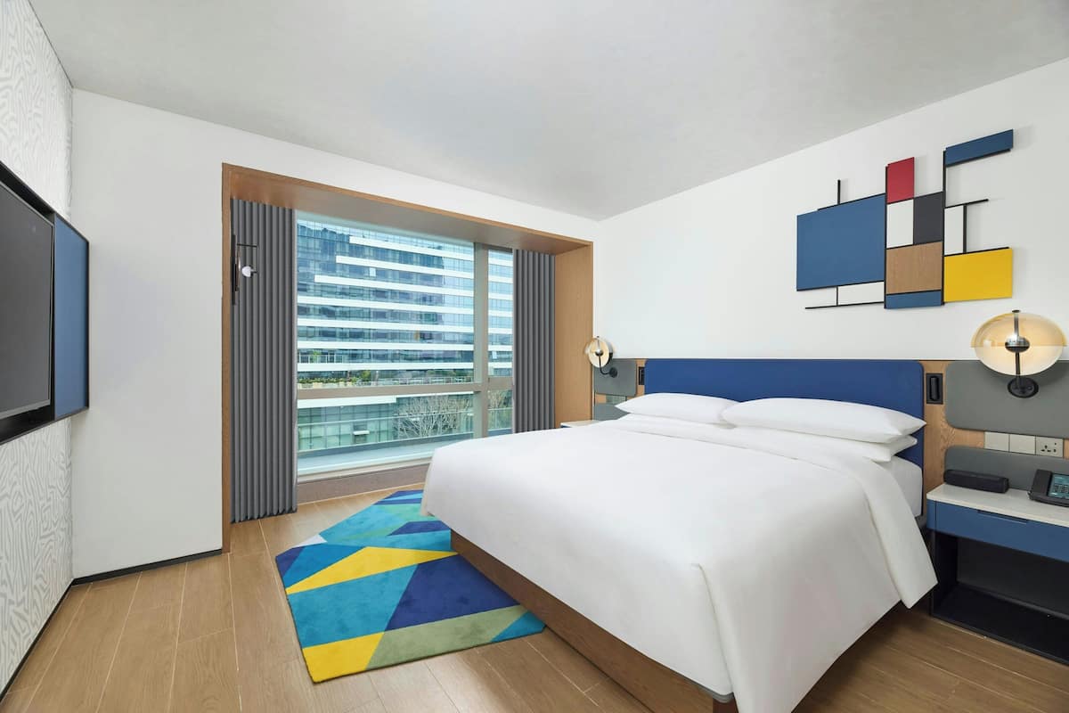 Quarto de hotel moderno com uma cama grande, televisão, mesa de cabeceira, abajur e vista para a cidade através de uma janela.