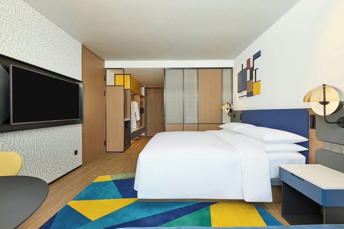 Quarto de hotel com cama, escrivaninha, televisão, guarda-roupa e banheiro visível por uma porta aberta.