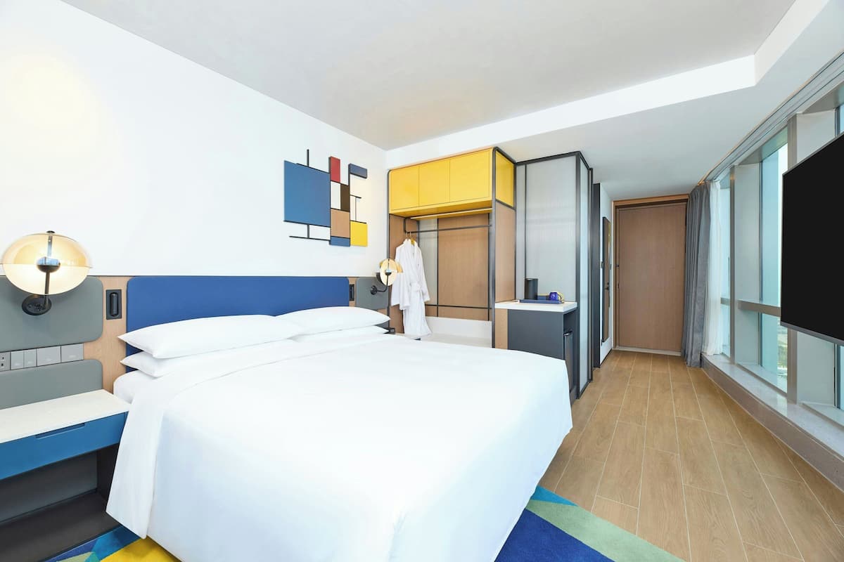 Quarto de hotel com cama, mesa de cabeceira, luminária, guarda-roupa e uma janela grande.
