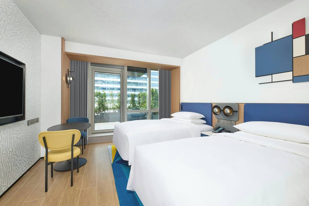 Quarto de hotel com duas camas, uma televisão, uma mesa de trabalho com cadeira e uma janela grande.