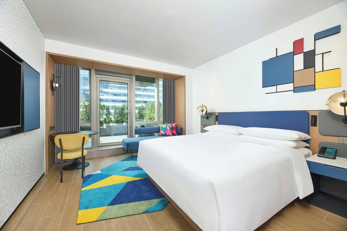 Quarto de hotel com cama, escrivaninha, cadeira, televisão e janela com vista para edifícios.