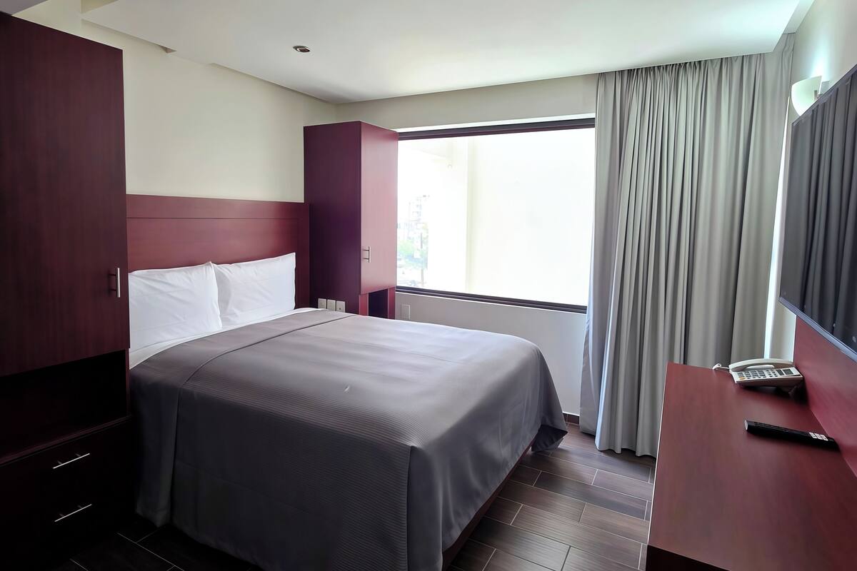 Quarto de hotel com uma cama grande, uma escrivaninha com telefone e uma janela com cortinas.