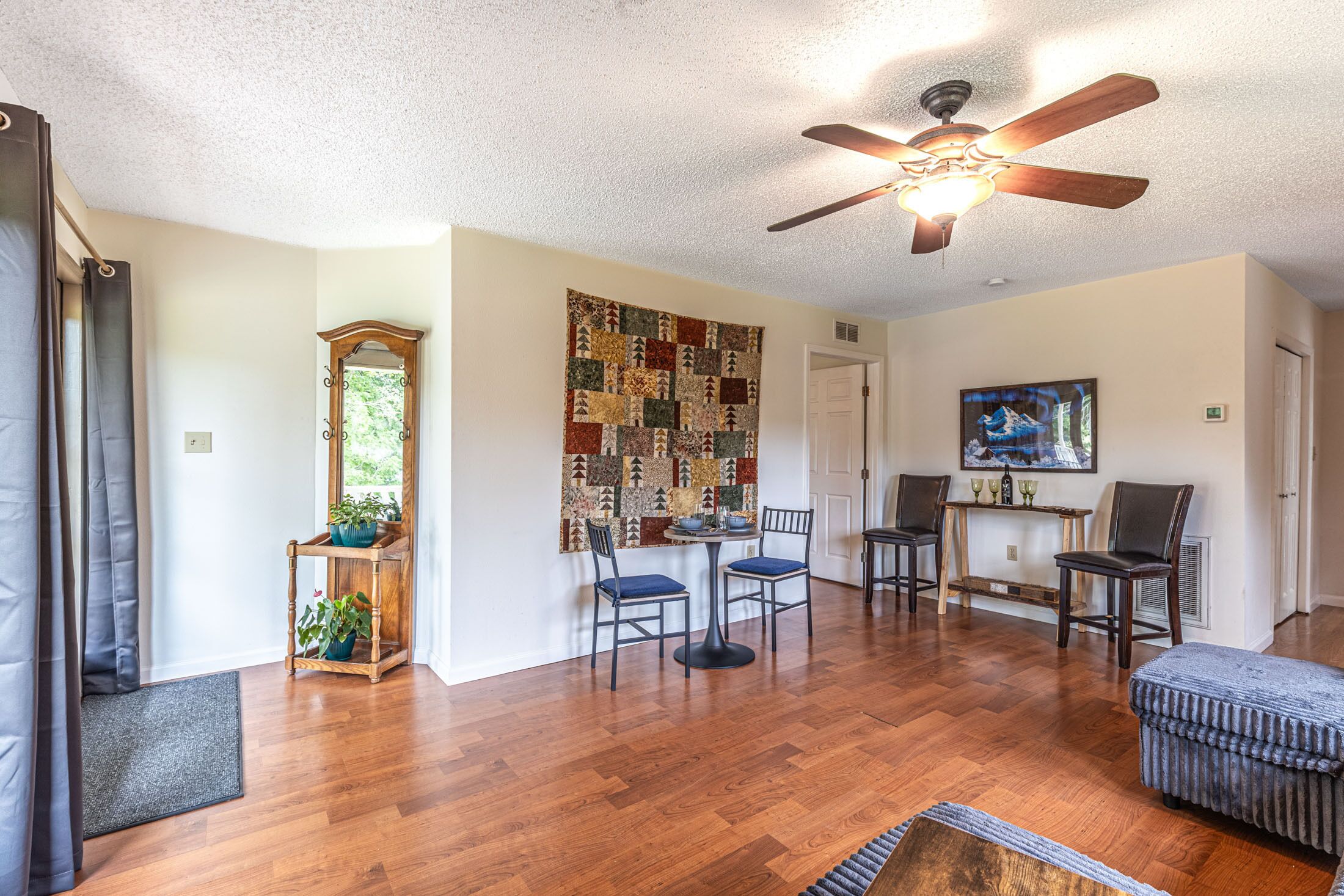 Um quarto amplo com piso de madeira, um ventilador de teto, uma grande tapeçaria na parede e um espaço de jantar com cadeiras e mesa.