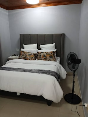 Quarto com cama, almofadas, mesa de cabeceira, ventilador e luminária no teto.