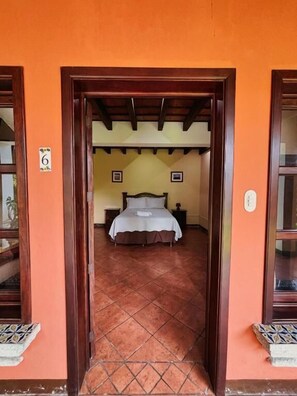 Uma porta dá acesso a um quarto com uma cama, uma mesinha e uma criadeira.