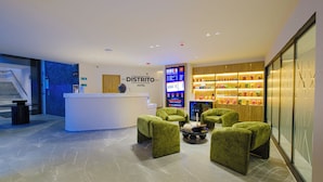 Área de recepção de hotel com balcão, poltronas verdes, tela digital e bar de lanches.