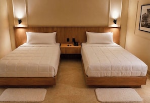Quarto de hotel com duas camas, cabeceiras de madeira, mesas de cabeceira e luminárias fixadas na parede.