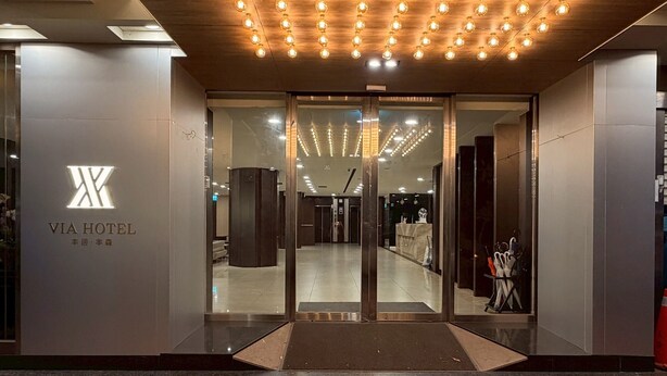 A entrada do VIA HOTEL, com design moderno, apresentando uma porta de vidro e um teto com várias luzes.