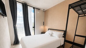 Quarto de hotel com uma cama, uma janela com cortinas, uma mesa de cabeceira e uma prateleira de metal preta.