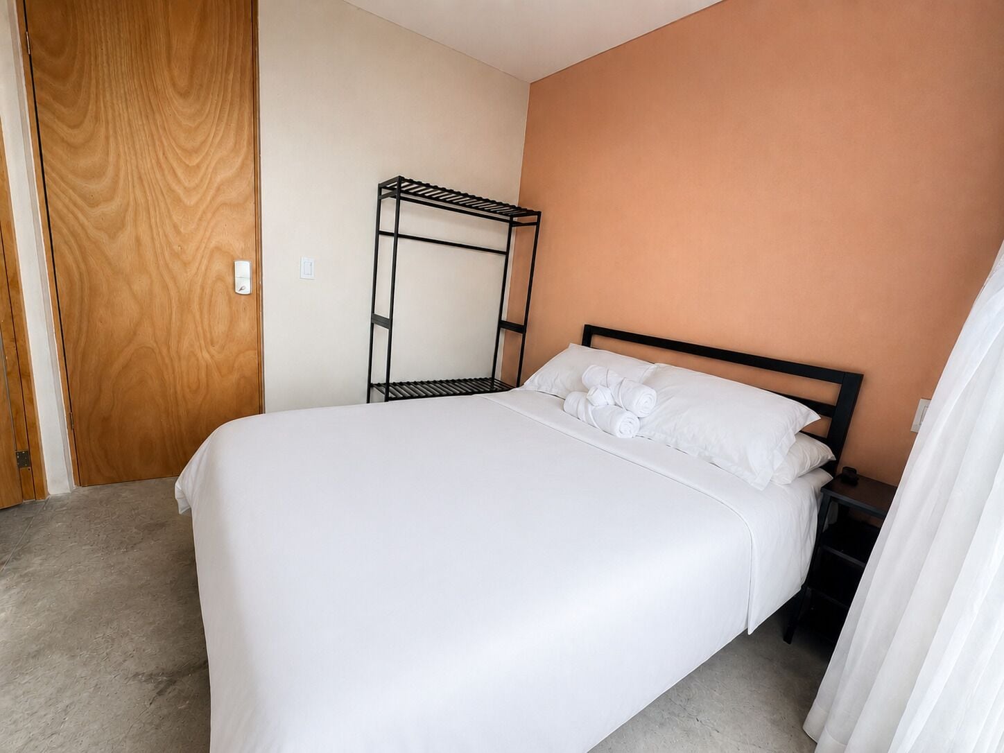 Um quarto com cama, porta de madeira, prateleira na parede e mesa de cabeceira.