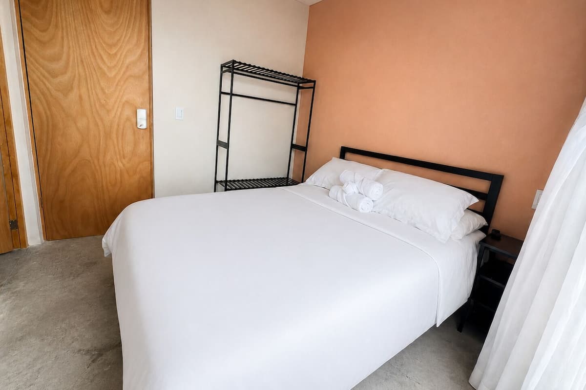 Um quarto com cama, porta de madeira, prateleira na parede e mesa de cabeceira.