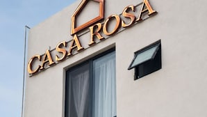 Um edifício com uma placa com o texto 'CASA ROSA' e duas janelas com cortinas.