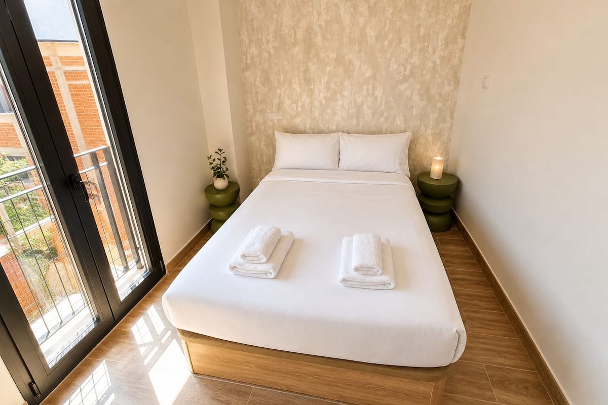 Quarto de hotel com uma cama, duas mesas de cabeceira, um vaso de planta e vista através de uma porta de vidro.