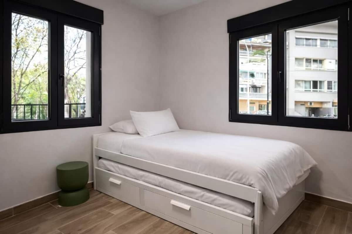 Quarto com cama, duas janelas, um banquinho verde e vista para edifícios.