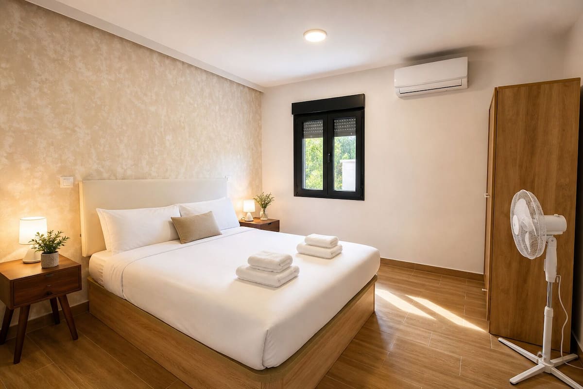 Quarto com cama, mesas de cabeceira, ventilador, ar condicionado e janela com persianas.