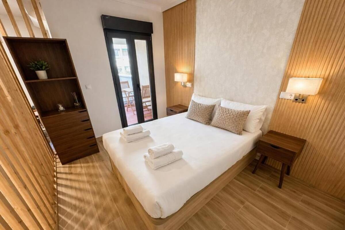 Quarto de hotel com uma cama grande, piso de madeira, uma prateleira de madeira com uma planta, uma porta que dá para uma varanda e duas luminárias fixadas na parede.