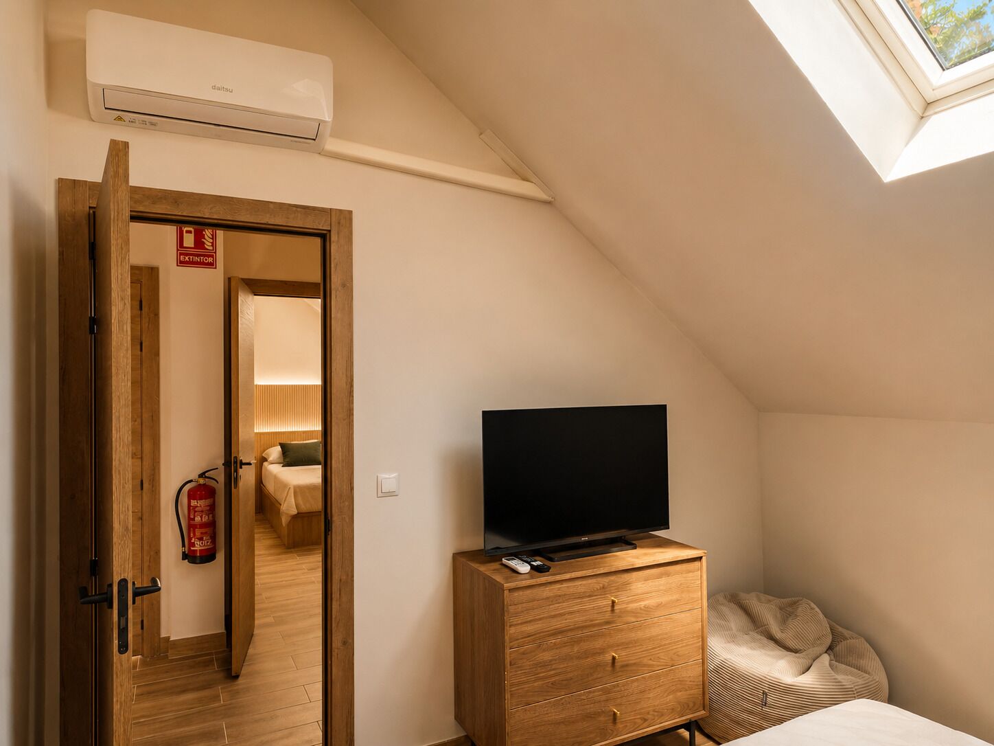 Um quarto com porta de madeira, televisão em uma cômoda de madeira, extintor de incêndio e claraboia.