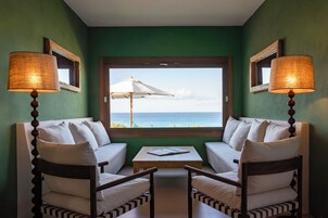 Quarto com dois sofás brancos, uma mesa de madeira e vista para o oceano através de uma janela com um guarda-sol.