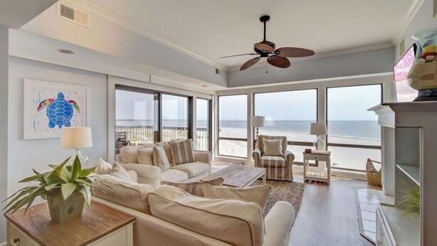 Uma sala de estar espaçosa com uma grande janela com vista para a praia, um ventilador de teto e um vaso de planta.