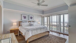 Quarto com cama, mesa de cabeceira, ventilador de teto e uma janela grande com persianas.