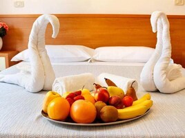 Quarto de hotel com cama, almofadas e uma cesta de frutas e toalhas na cama.