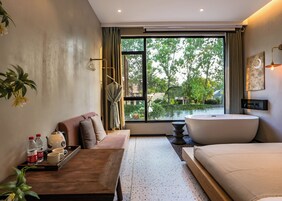 Quarto de hotel moderno com uma grande janela, banheira, sofá, mesa de madeira com garrafas e bandeja, e uma planta em vaso.