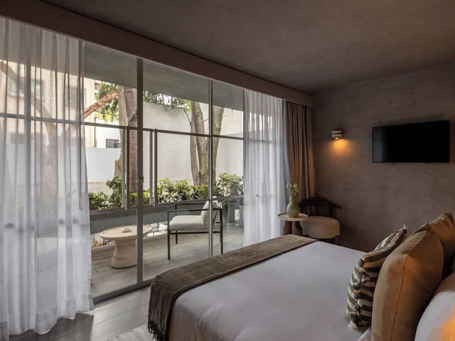 Quarto com uma janela grande, uma cama, televisão na parede e uma pequena mesa com cadeira do lado de fora.
