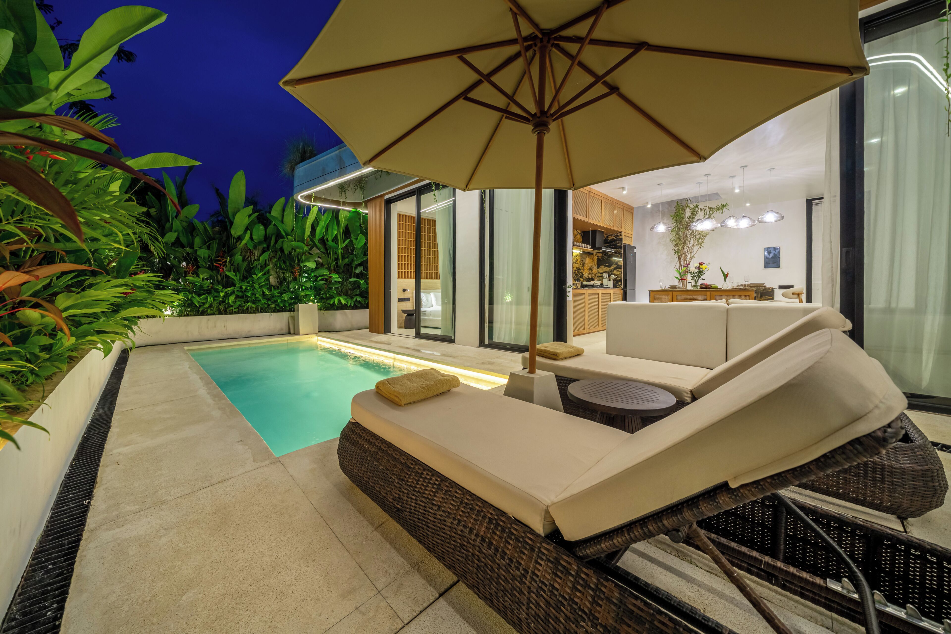 Uma área moderna de piscina externa com espreguiçadeira, guarda-sol e espaço de refeições visível pelas portas de vidro.