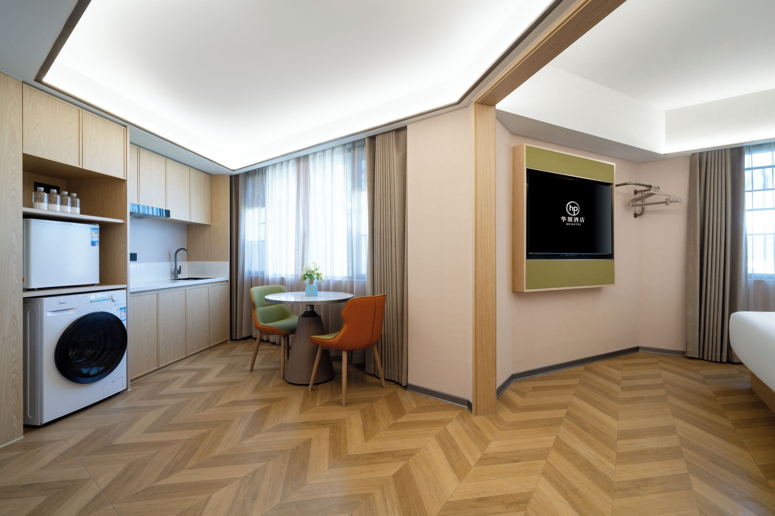 Quarto de hotel moderno com cozinha, mesa de jantar com cadeiras, TV fixada na parede e uma grande janela com cortinas.