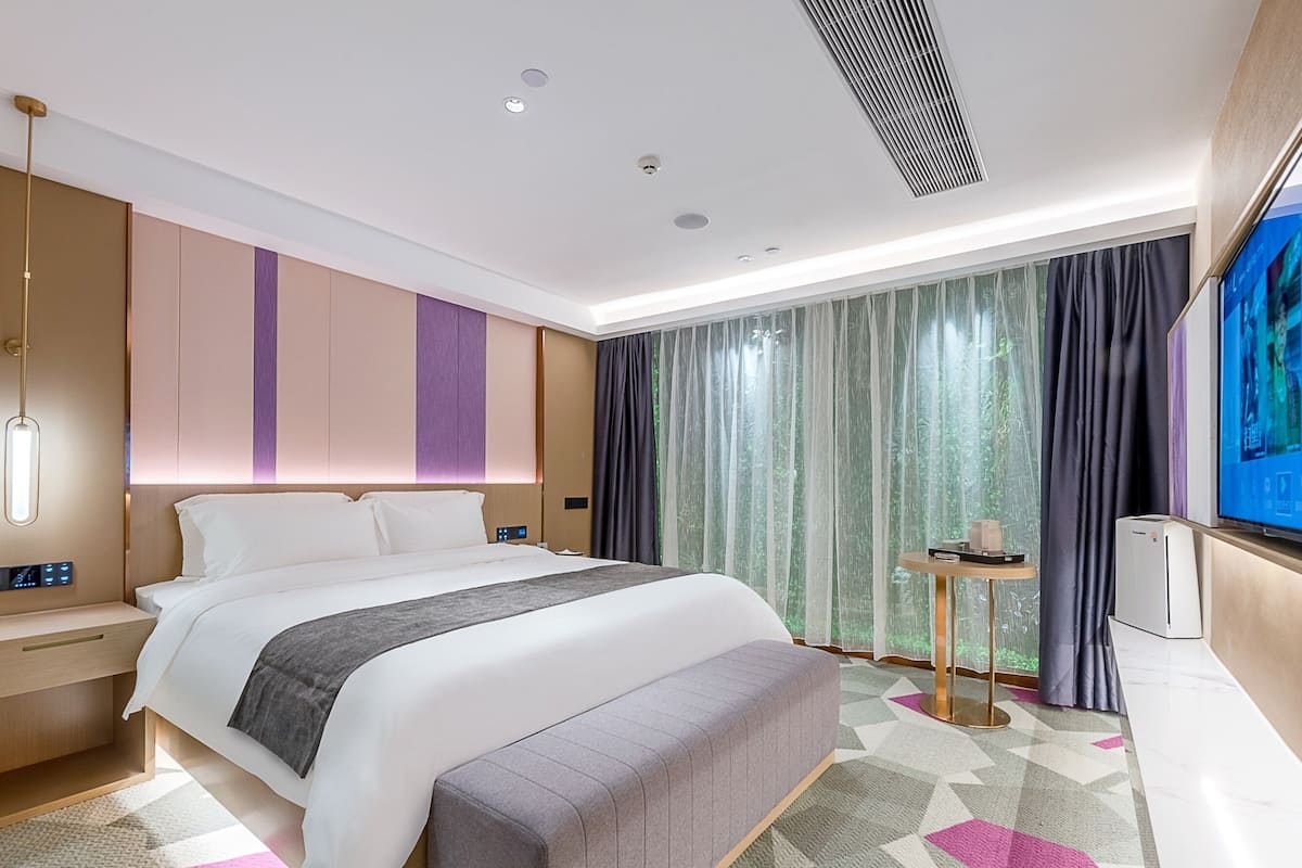 Quarto de hotel moderno com uma cama grande, mesas de cabeceira, uma TV e vista para o verde através de cortinas leves.
