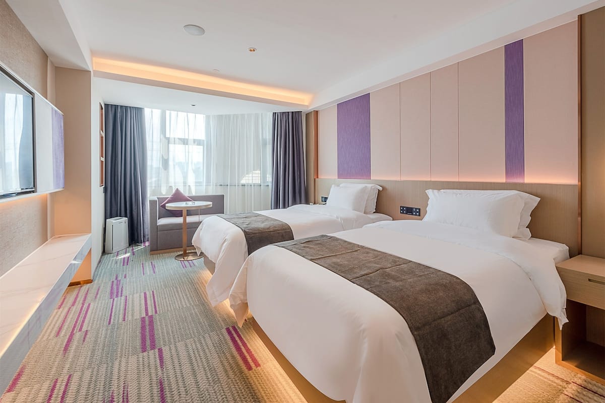 Quarto de hotel com duas camas, uma mesa pequena, uma cadeira e uma janela grande com cortinas.