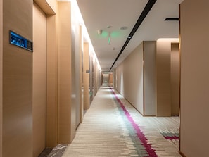 Um corredor de hotel comprido e estreito, com carpete estampado, paredes de madeira e um relógio digital à esquerda.